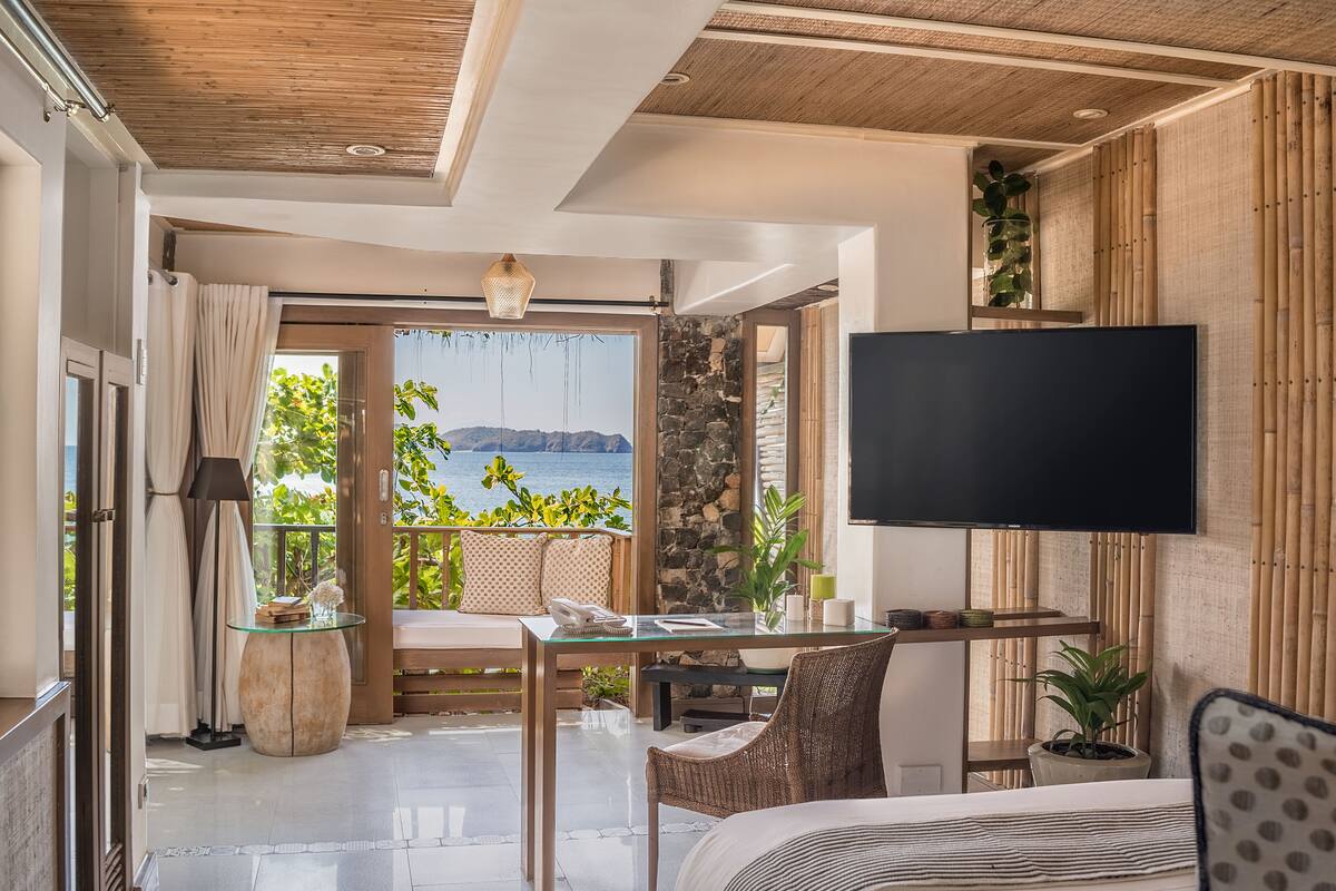 Quarto moderno com cama, mesa de vidro, televisão e vista para o mar.