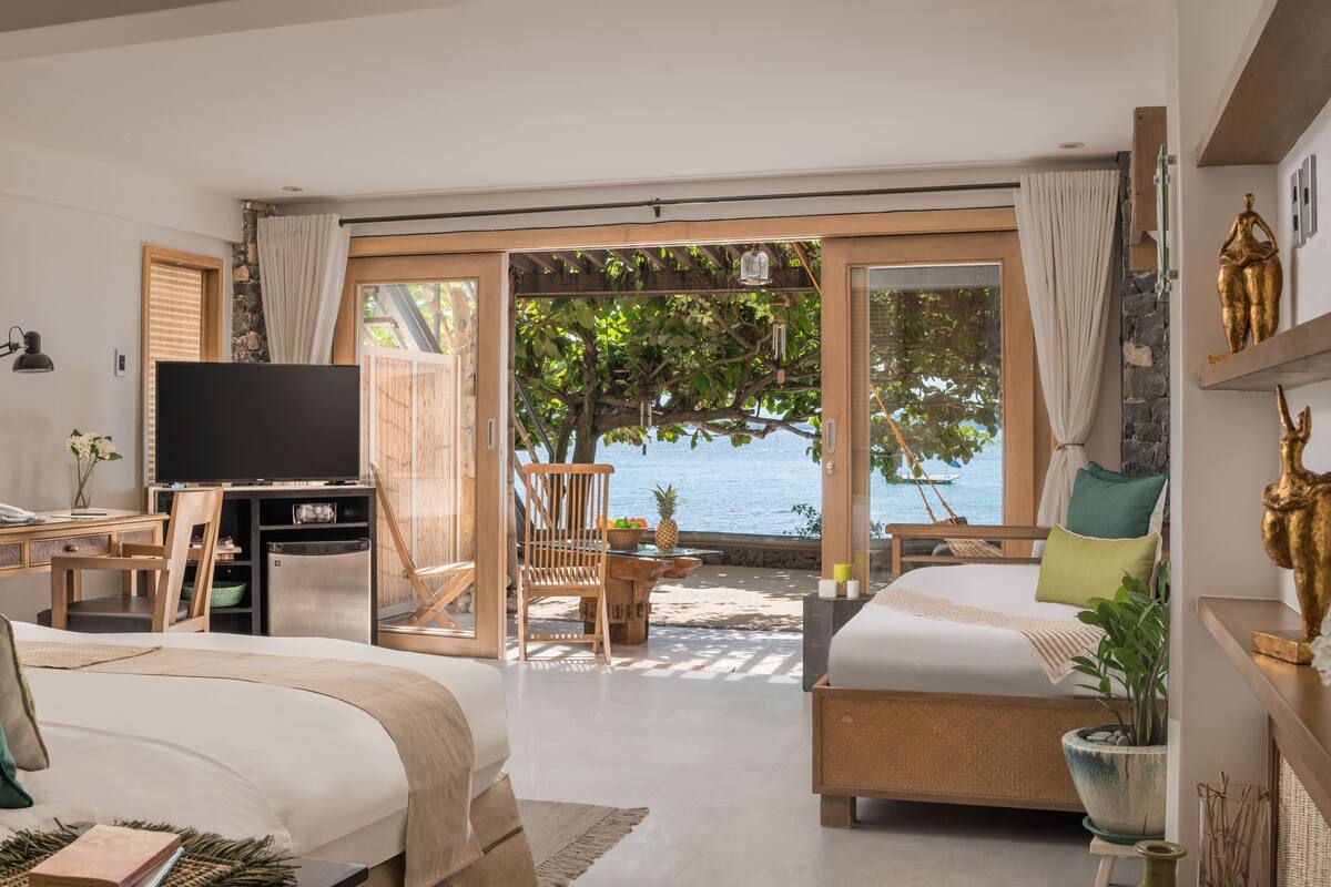 Quarto com duas camas, uma escrivaninha, televisão e vista para o mar.