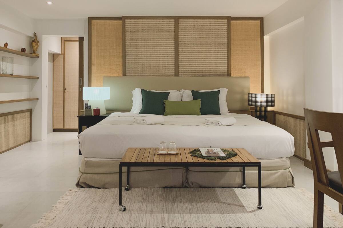 Quarto de hotel bem arrumado com uma cama grande, uma mesa de cabeceira com abajur, uma pequena mesa com um copo de água e uma cadeira.