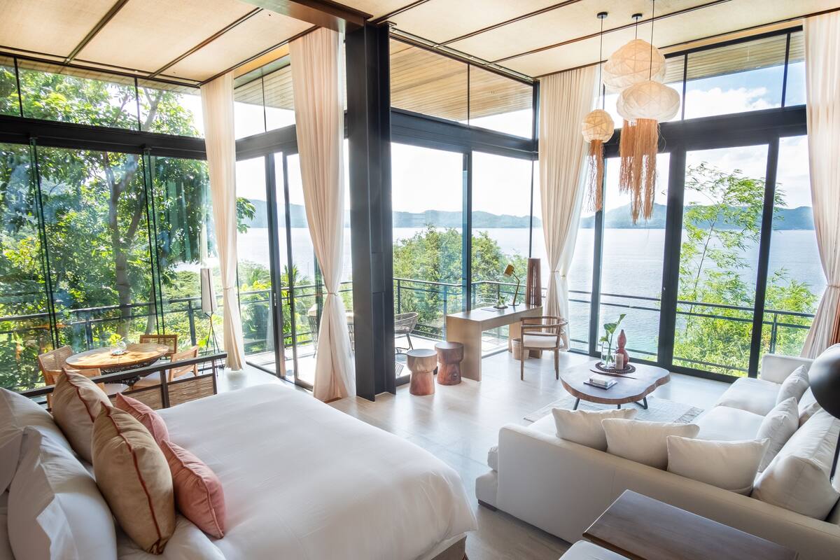 Quarto de hotel moderno com cama grande, sofá, mesa de jantar e vista para um lago.