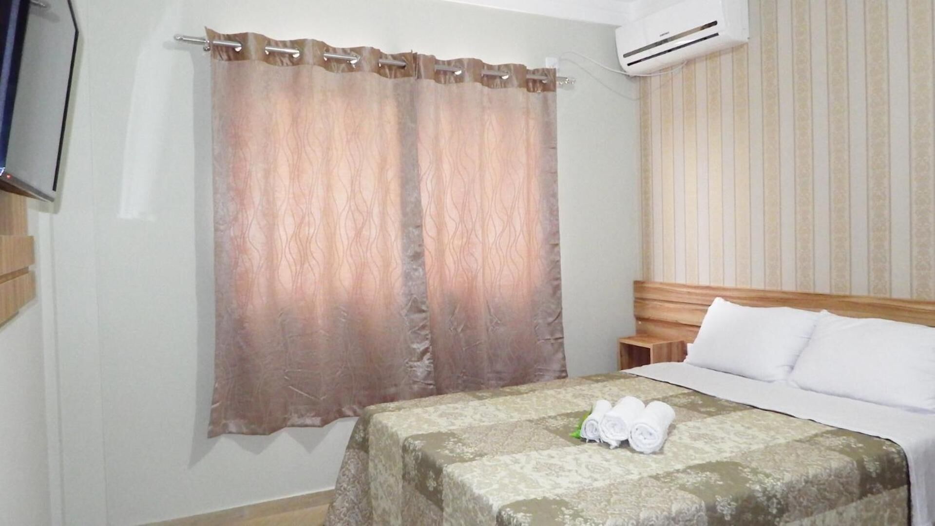 Quarto de hotel com uma cama, mesa de cabeceira de madeira, televisão e ar condicionado.
