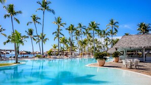 Piscina de resort com palmeiras, espreguiçadeiras e um edifício com telhado de palha.