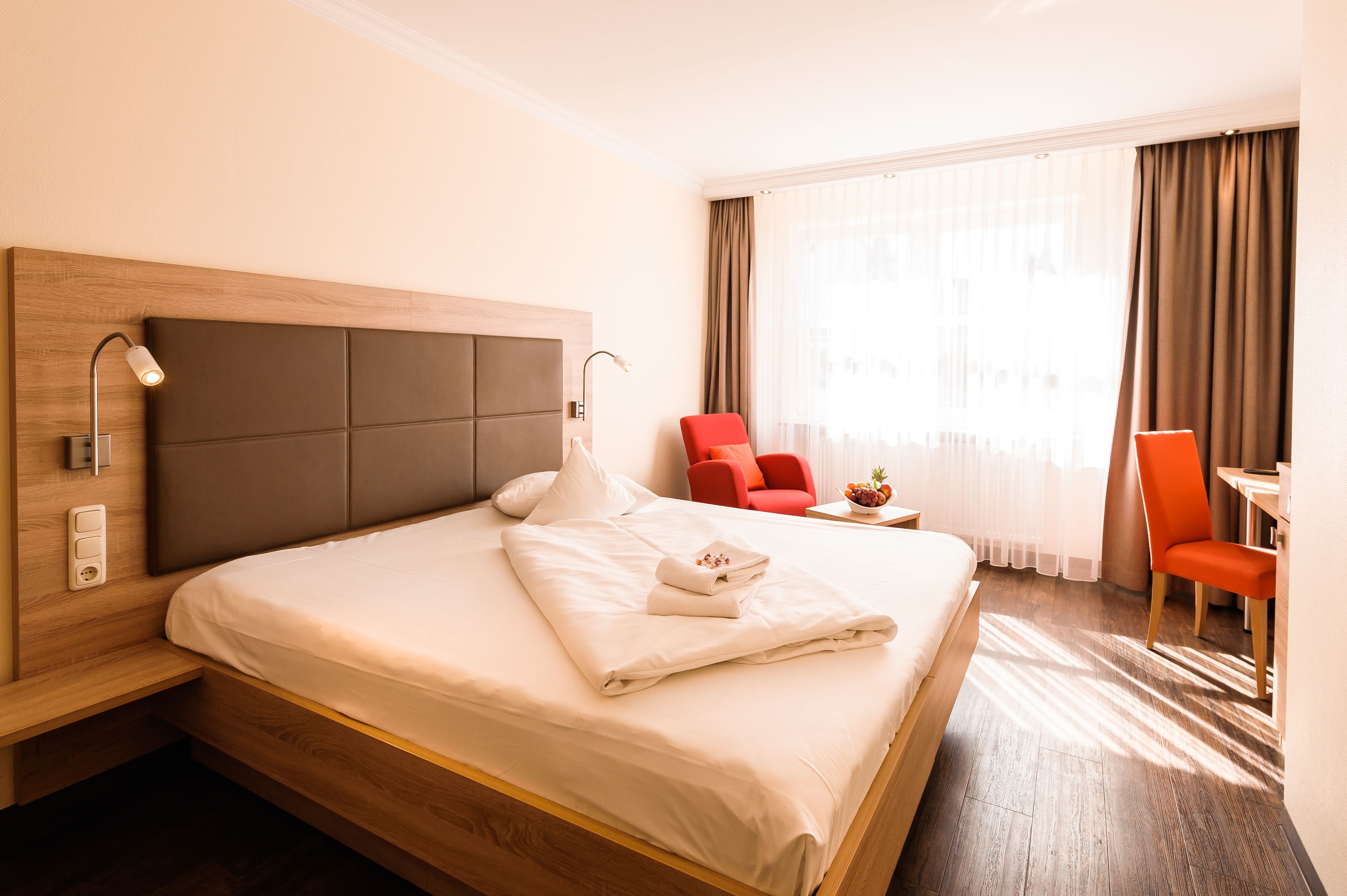 Quarto de hotel com cama, escrivaninha, cadeira, luminária e janela com cortinas.