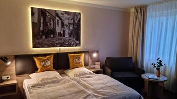 Quarto de hotel com cama, duas almofadas, mesa de cabeceira, cadeira, mesinha e janela com cortinas.
