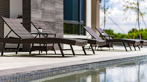 Uma área na beira da piscina com espreguiçadeiras e uma mesa, piscina com azulejos e um edifício com janelas amplas e fachada de pedra.