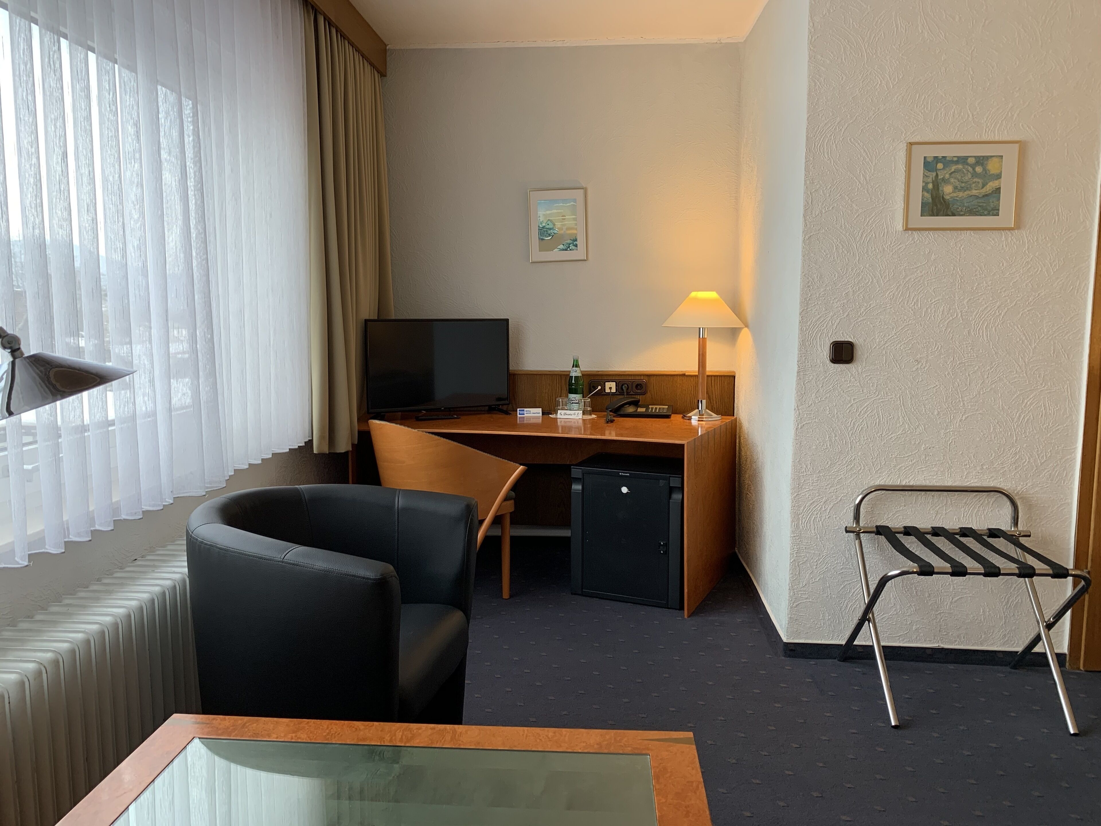 Quarto de hotel com escrivaninha, cadeira, televisão, luminária, radiador e um pequeno frigorífico.