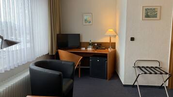 Quarto de hotel com escrivaninha, cadeira, televisão, luminária, radiador e um pequeno frigorífico.
