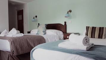 Quarto de hotel com duas camas, cada uma com cabeceira de madeira, roupa de cama branca e toalhas dobradas. Há duas luminárias fixadas na parede e uma porta que dá para outro quarto.