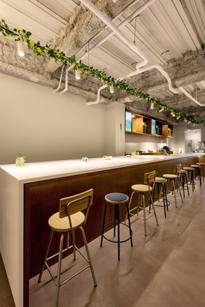 Uma área de bar moderna com um balcão comprido, banquetas de madeira e teto com canos aparentes e vegetação.