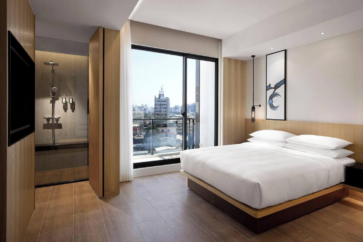Quarto de hotel moderno com uma cama grande, chuveiro, vista para a cidade e televisão montada.