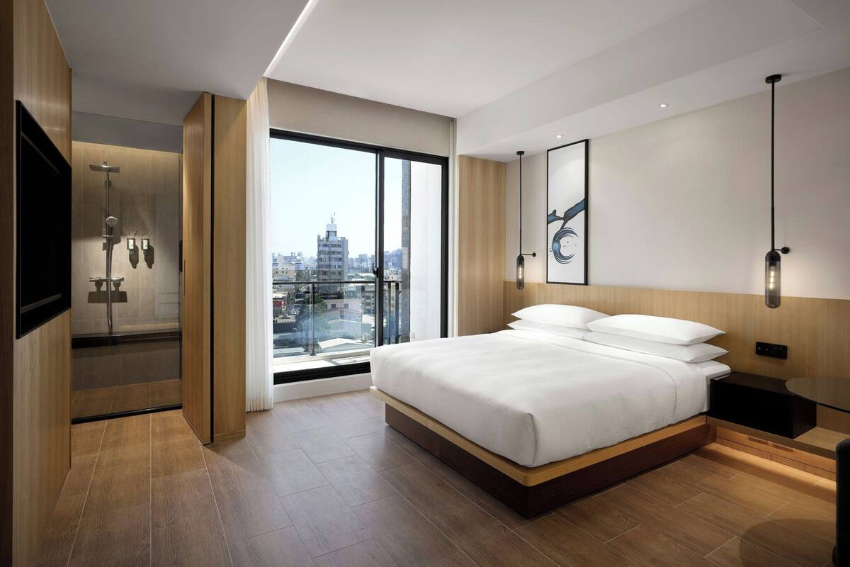 Quarto de hotel moderno com uma cama grande, chuveiro, vista da cidade e TV na parede.