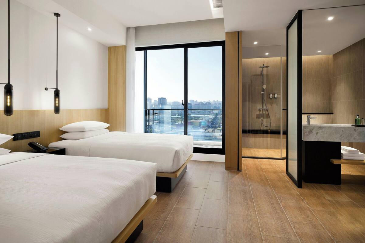 Quarto de hotel com duas camas, uma janela grande com vista para a cidade, chuveiro e banheiro com bancada de mármore.