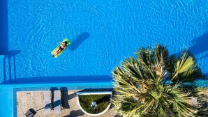 Piscina azul límpida com uma pessoa em um tapete flutuante, cercada por palmeiras e espreguiçadeiras.