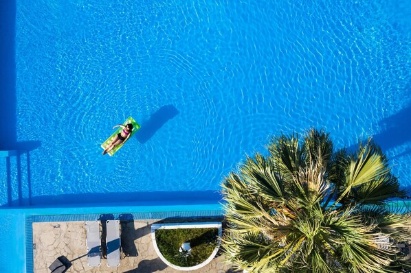 Piscina azul límpida com uma pessoa em um tapete flutuante, cercada por palmeiras e espreguiçadeiras.