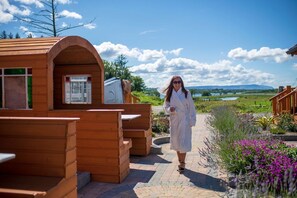 Uma cabana de madeira com varanda coberta, um banco e um pequeno jardim com flores roxas.