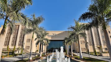 Exterior do Triumph Hotel & Conference Center, com uma fonte central, palmeiras e um céu azul límpido.