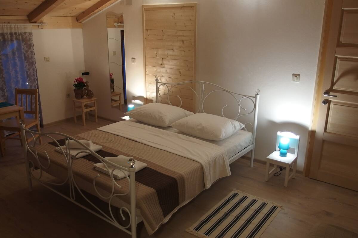 Quarto com estrutura de cama de metal, cabeceira de madeira, mesas de cabeceira com abajures, piso de madeira e uma janela com cortinas.