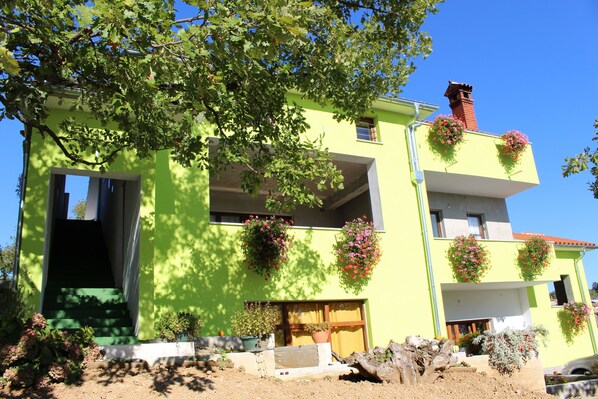 Um edifício de dois andares com fachada verde, uma escadaria visível e vasos de flores pendurados nas varandas.