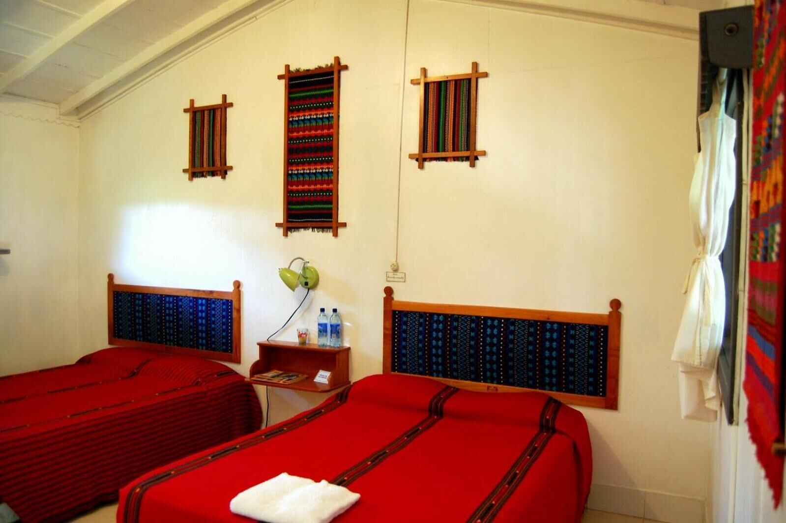 Quarto com duas camas, cada uma com cabeceira de madeira e roupa de cama vermelha. Há uma pequena mesa com abajur e uma garrafa de água.