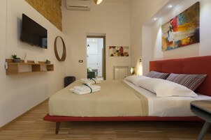 Quarto de hotel com uma cama grande, televisão de tela plana, prateleira de madeira com produtos de higiene pessoal, um quadro e cabeceira vermelha.
