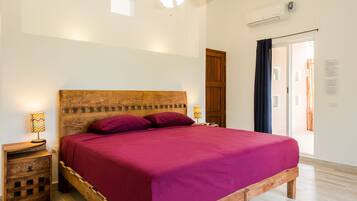 Quarto com estrutura de cama de madeira, colcha vermelha, duas mesas de cabeceira com luminárias, ventilador de teto e porta de vidro corrediça para área externa.