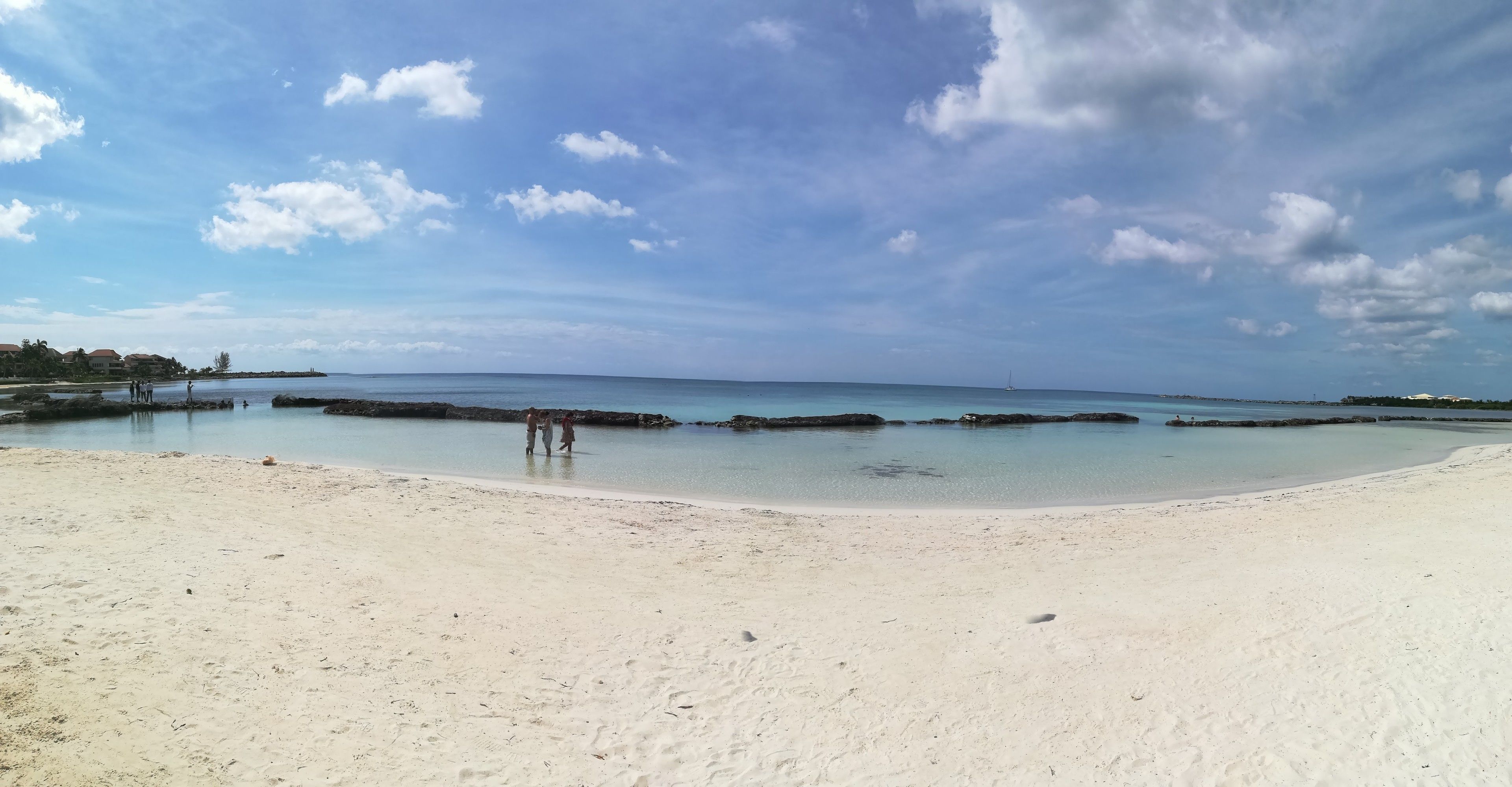 Uma praia com águas cristalinas, algumas pessoas e um veleiro ao longe.