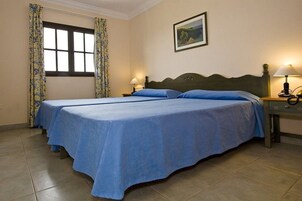 Quarto de hotel com duas camas cobertas por lençóis azuis, uma janela com cortinas, um quadro na parede e duas luminárias de cabeceira.