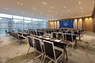 Uma sala de conferências com fileiras de mesas e cadeiras, uma tela grande exibindo o logotipo da Hilton e amplas janelas com vista para a cidade.