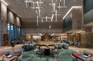 Um amplo hall de hotel com área de bar central, vários espaços de estar e teto alto com luminárias modernas.