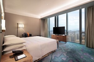 Quarto de hotel com uma cama grande, televisão de tela plana, escrivaninha com abajur e vista da cidade através de janelas amplas.