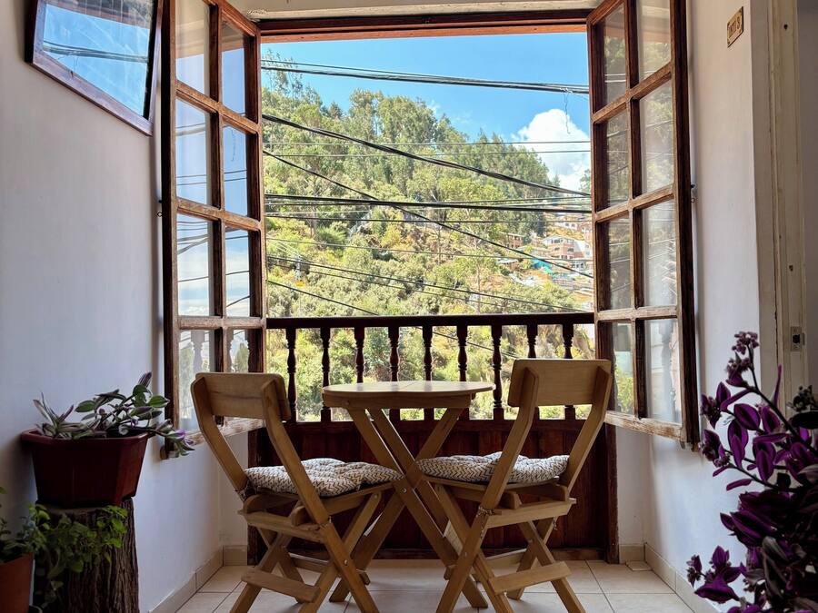 Uma varanda com cadeiras e mesa de madeira, plantas em vasos e vista para uma encosta.