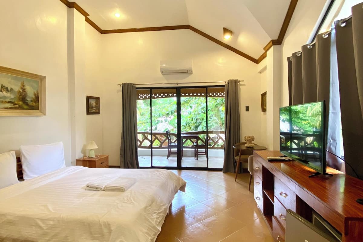 Quarto de hotel com cama, escrivaninha com televisão, ventoinha no teto e vista para o exterior.