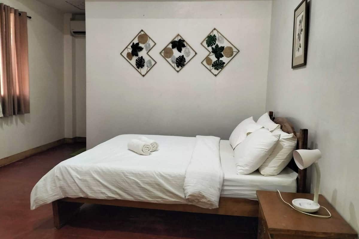 Cama bem arrumada com roupa de cama e almofadas brancas, mesa de cabeceira com abajur e três objetos decorativos fixados na parede.