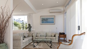 Uma sala de estar com sofá, mesa de centro de vidro, cadeira de madeira, um quadro e vista para o exterior.
