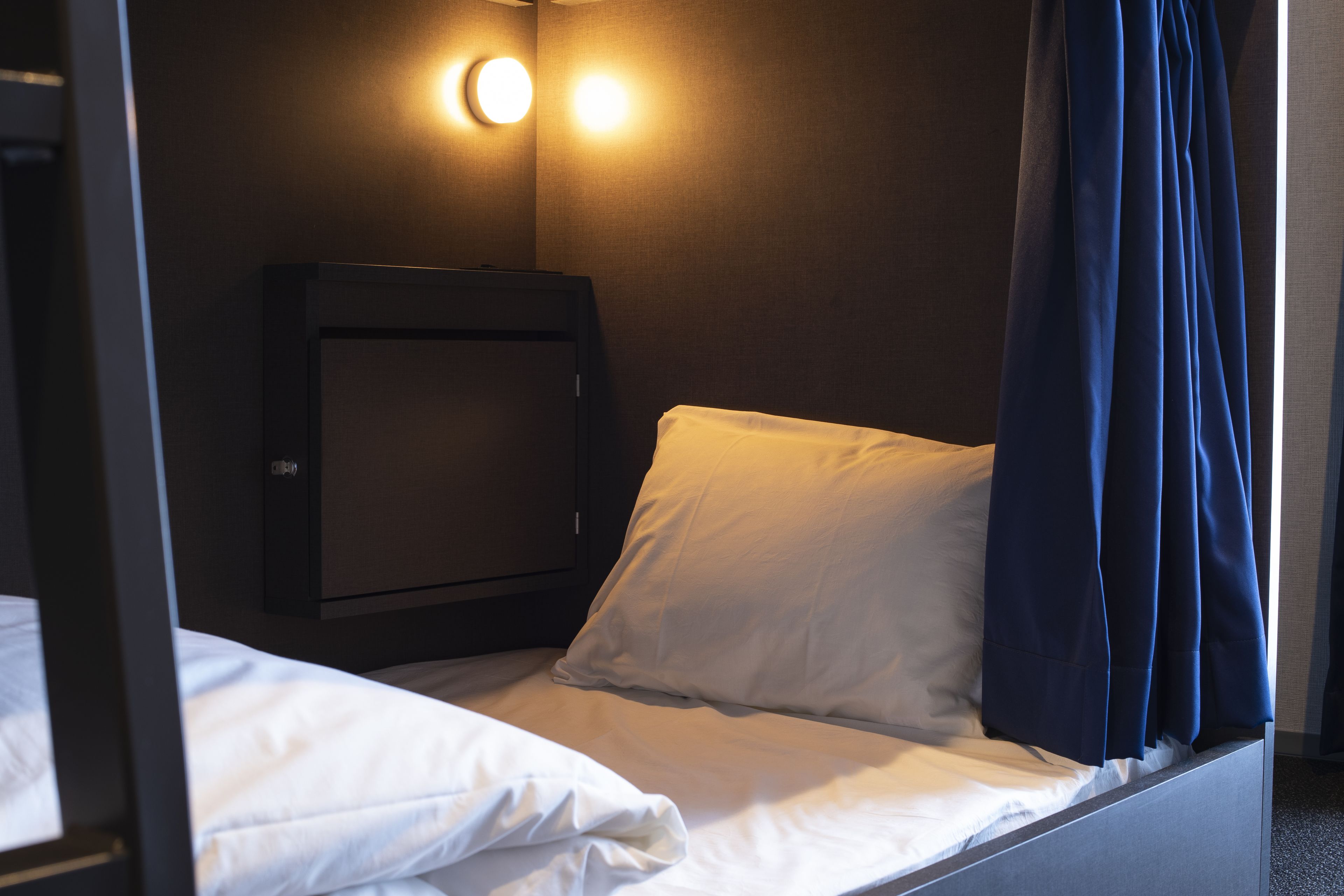 Uma cama de solteiro com roupa de cama branca, cabeceira escura e luminária fixada na parede.
