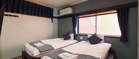 Quarto de hotel com duas camas, uma janela com cortinas, ar condicionado e piso de madeira.
