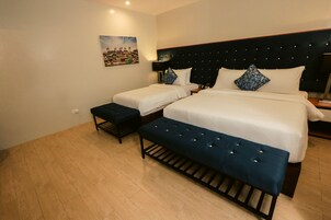 Quarto de hotel com duas camas, um banquinho, uma mesinha de cabeceira e uma luminária na parede.