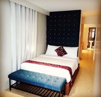 Quarto de hotel com uma cama grande, um banquinho, cabeceira escura e vista para o banheiro.