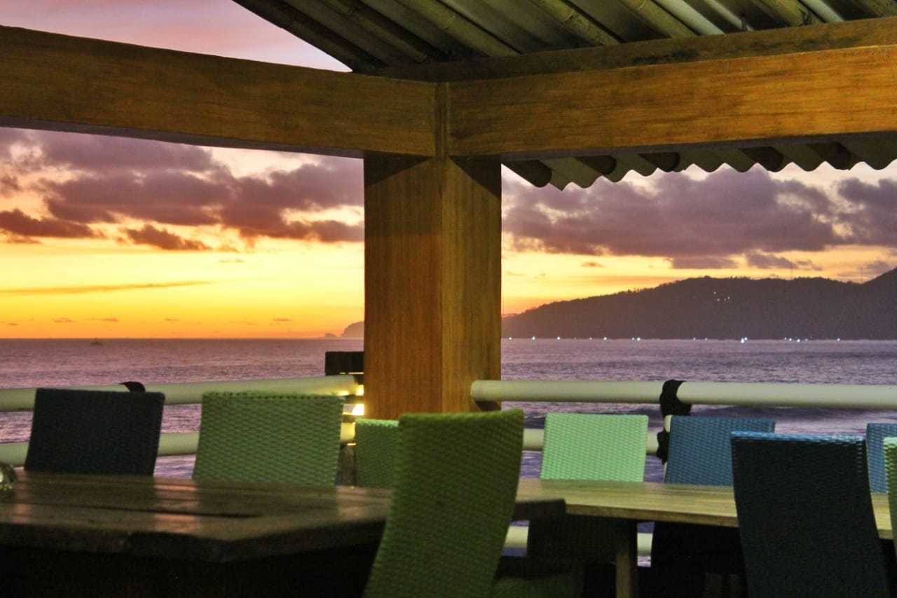 Um restaurante à beira-mar com mesas ao ar livre e vista para o oceano ao pôr do sol.