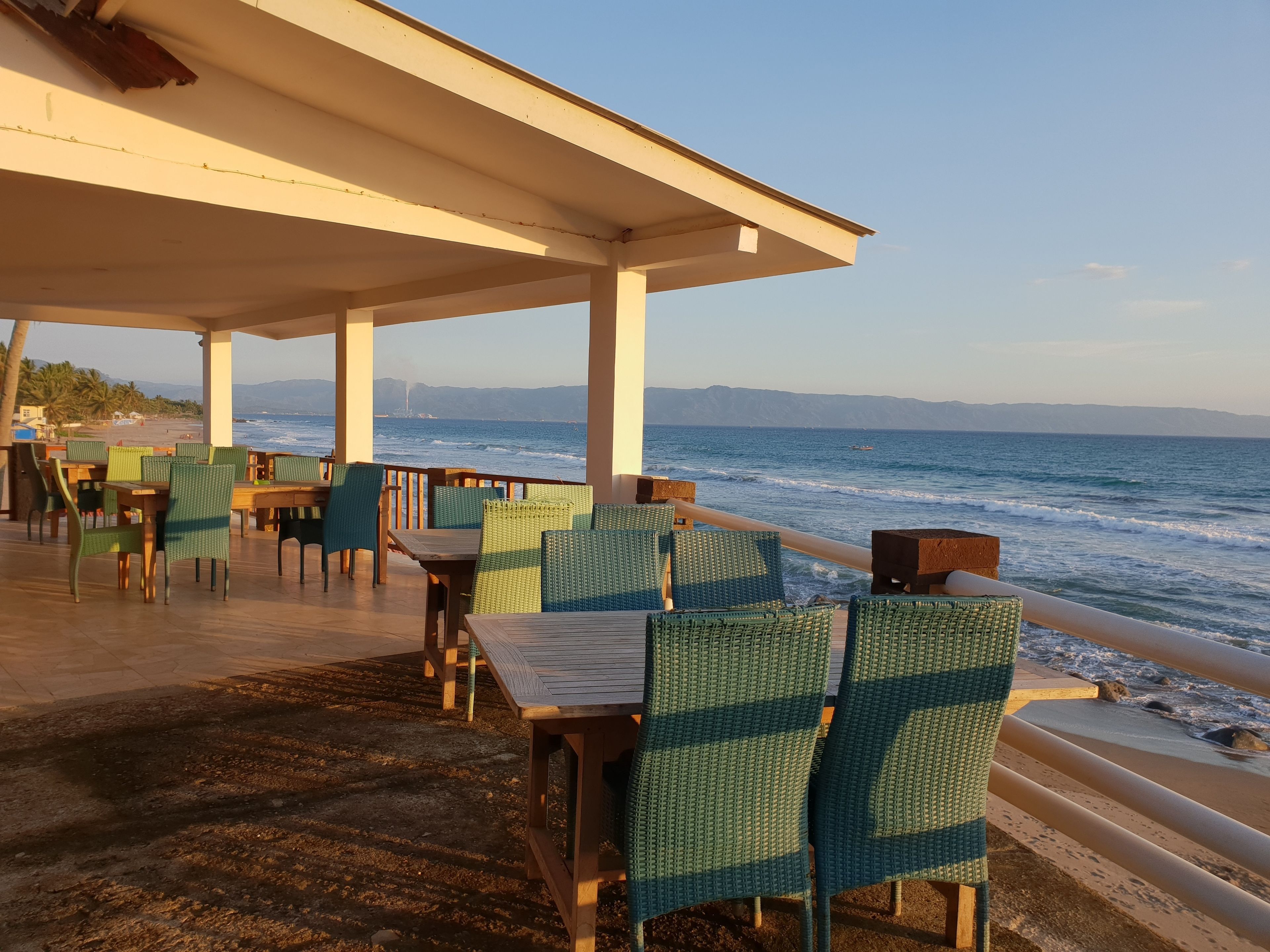 Um restaurante à beira-mar com mesas ao ar livre, vista para o oceano e montanhas ao longe.