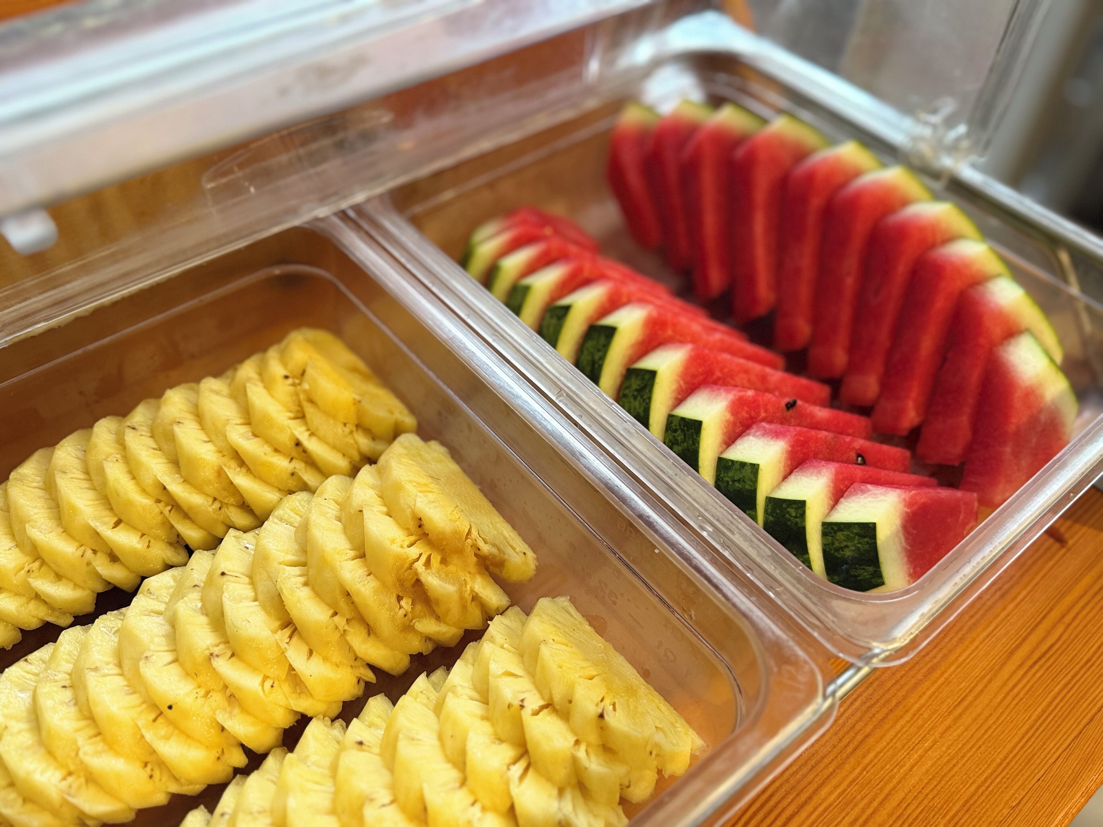 Exposição de frutas com abacaxi e melancia fatiados em um recipiente de plástico transparente.