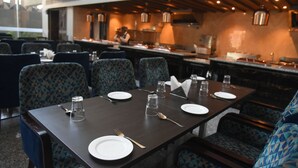 Restaurante com mesas preparadas para refeições, cadeiras com estofamento estampado e área de cozinha ao fundo.