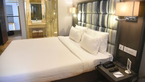 Quarto de hotel com uma cama grande, uma mesa de cabeceira com telefone e computador, um espelho e banheiro com box de vidro.