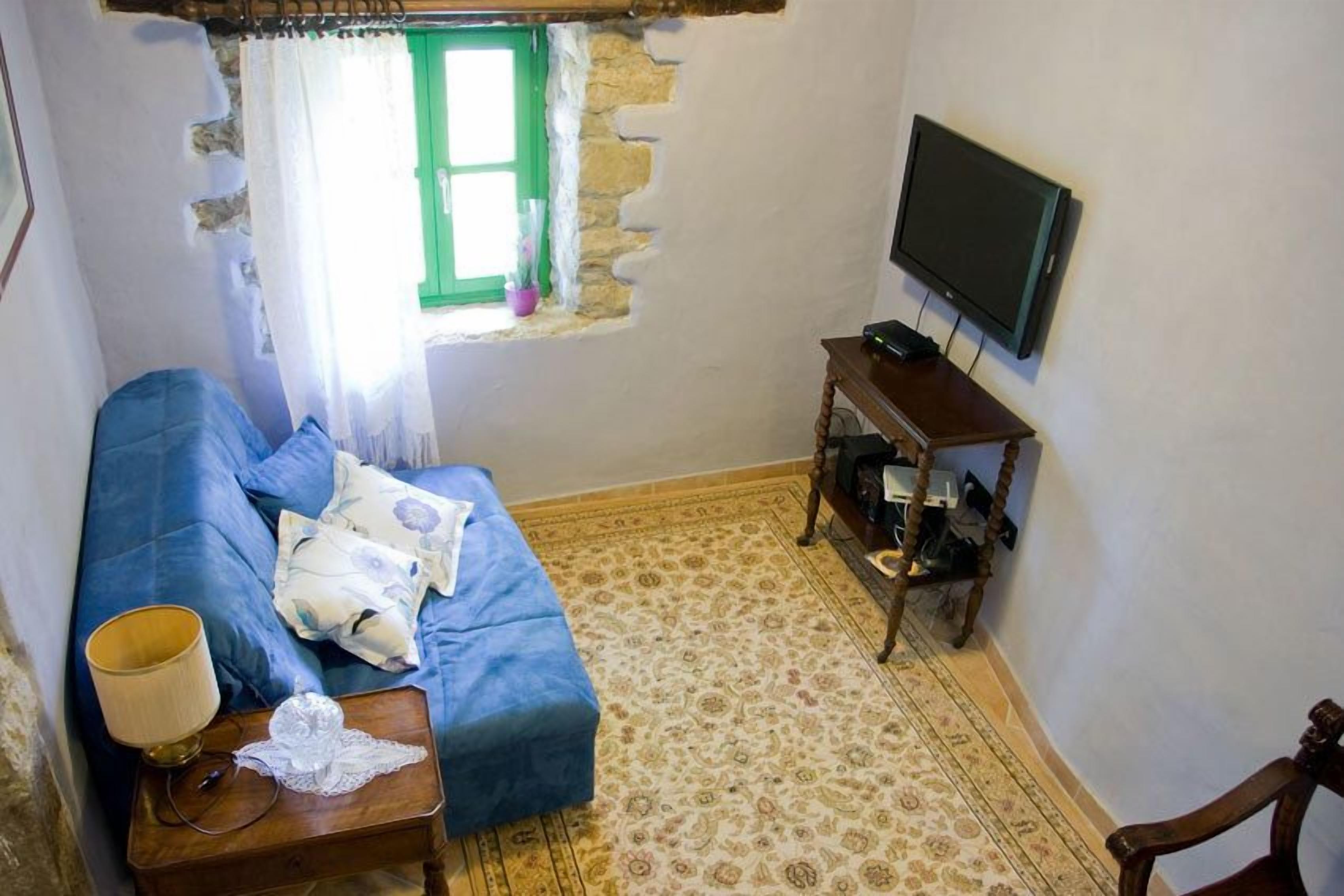 Um quarto aconchegante com um sofá azul, uma pequena mesa de madeira com uma luminária, uma TV sobre um suporte de madeira e um tapete estampado.