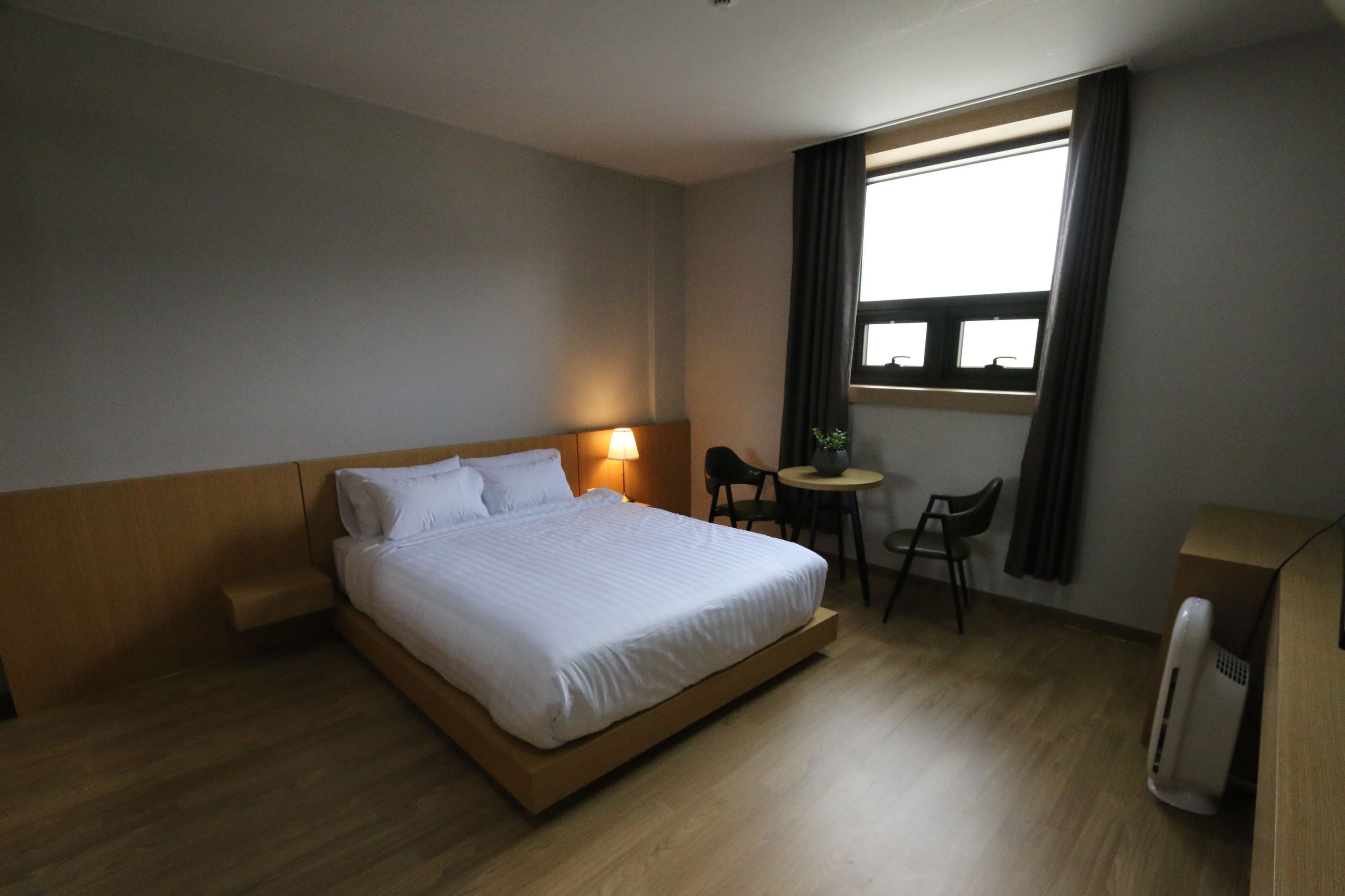 Quarto de hotel com cama, mesa de cabeceira, luminária, pequena mesa com cadeiras e janela com cortinas.