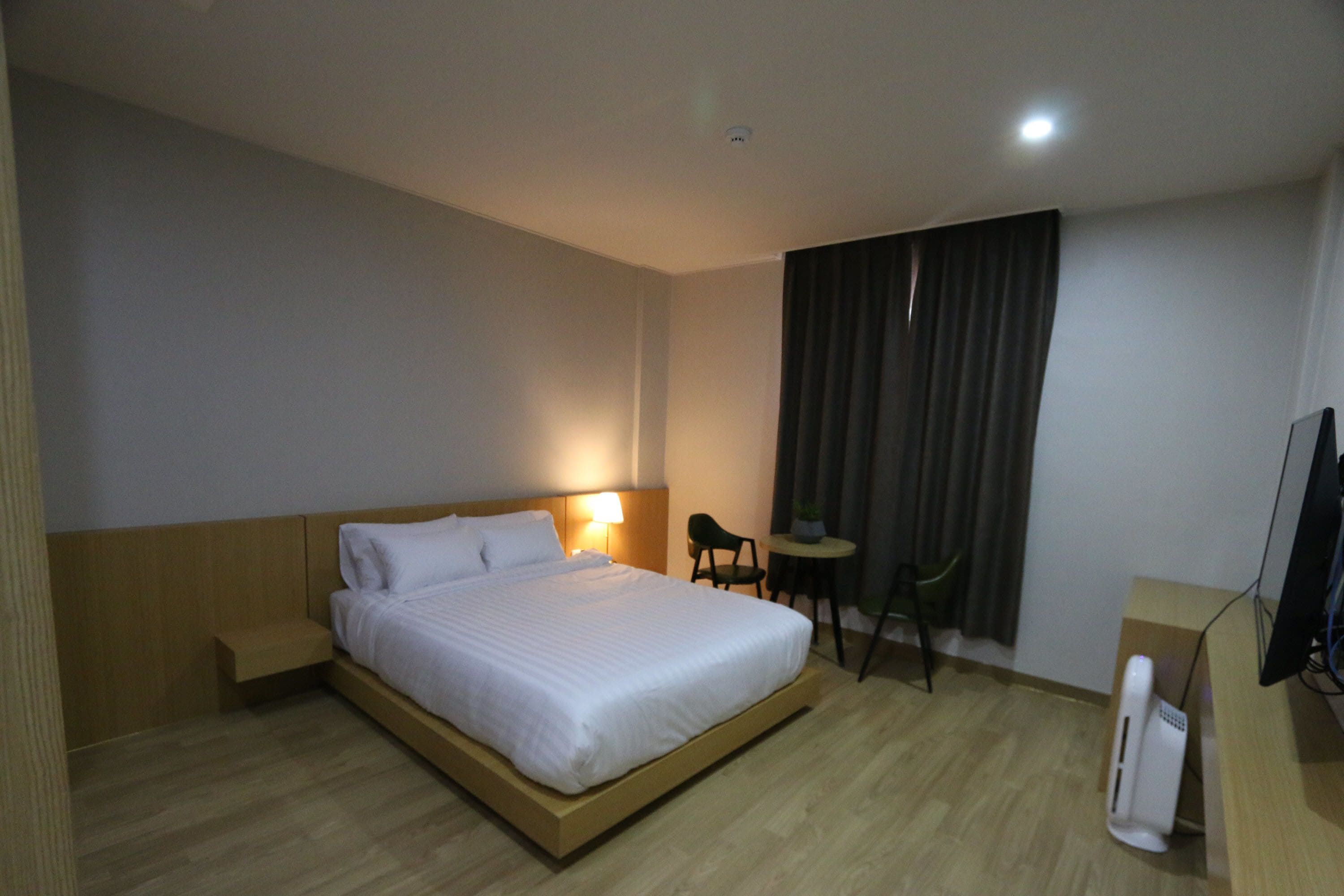 Quarto de hotel com cama de madeira, uma escrivaninha, uma cadeira, televisão e janela com cortinas.