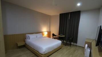 Quarto de hotel com cama de madeira, uma escrivaninha, uma cadeira, televisão e janela com cortinas.