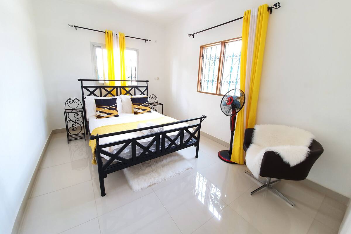 Um quarto com cama, cadeira, ventoinha e janela com cortinas.