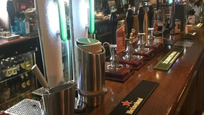 Uma área de bar com várias torneiras de cerveja, um registo de caixa e uma variedade de garrafas e copos em prateleiras.