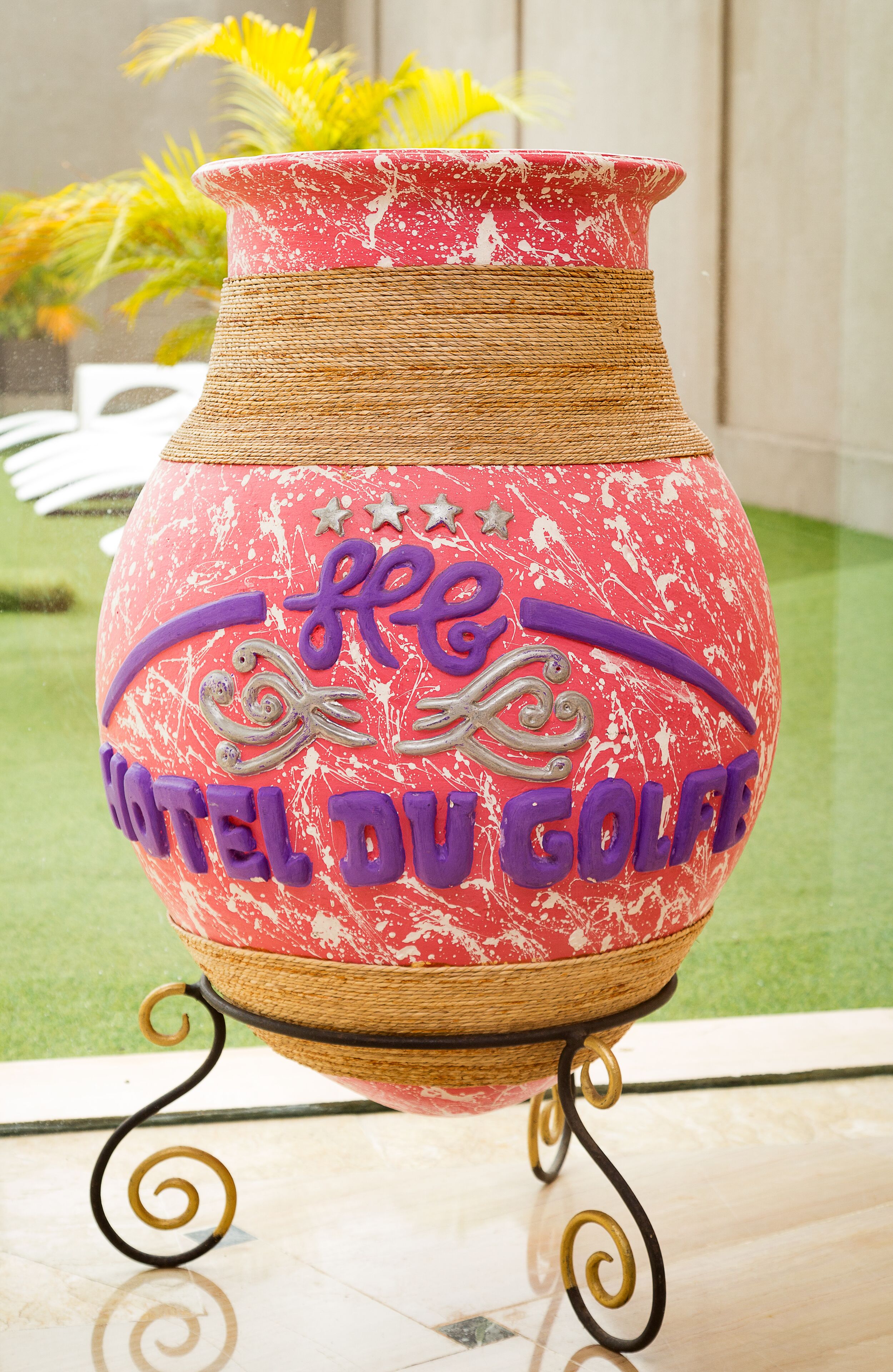 Um vaso vermelho com a inscrição "HOTEL DU GOLF", colocado sobre um suporte.